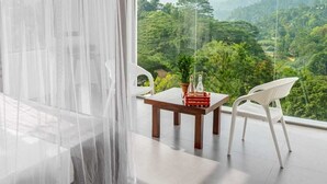 Quarto com vista para uma paisagem verde exuberante, uma mesa de madeira com garrafas e cadeiras brancas.