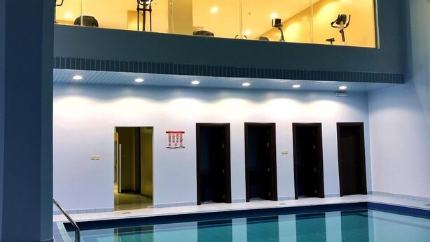 Uma piscina interior moderna com fundo de vidro transparente, um segundo nível com assentos e iluminação, e chão revestido de azulejos.