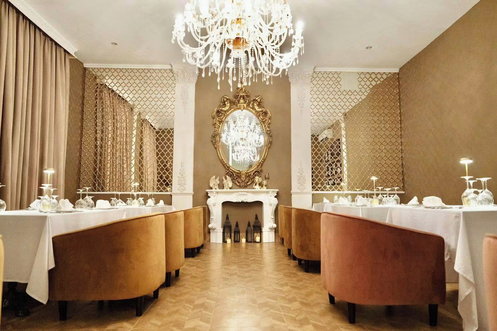 Uma sala de jantar formal com lareira central, espelhos ornamentados e um candelabro. As mesas estão postas com toalhas brancas e copos.