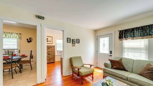 Uma sala de estar com sofá, uma mesa de jantar com cadeiras, uma cadeira de madeira, uma mesa de centro com uma planta e uma porta com janela.