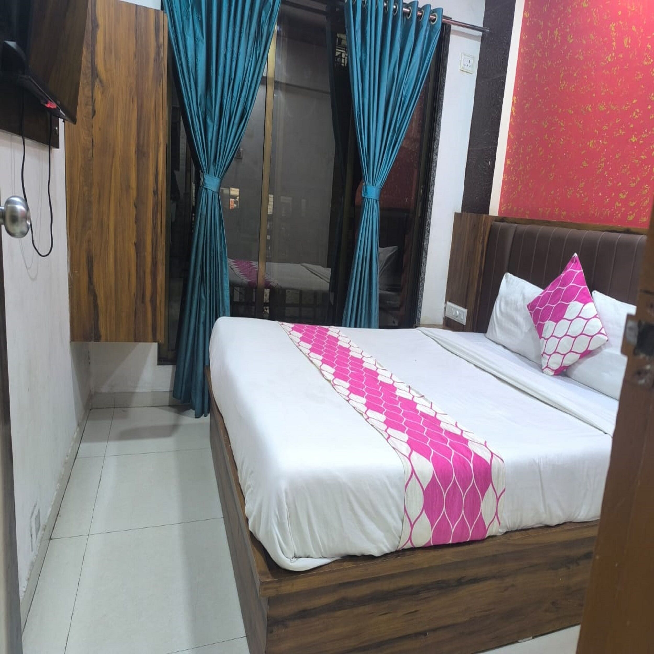 Quarto com cabeceira de madeira, cama com roupa de cama branca e manta estampada em rosa e branco, cortinas verde-água e parede vermelha com detalhes dourados.