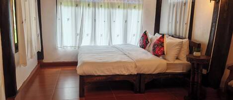 Quarto com cama de madeira, roupa de cama branca, tapete floral e janela com cortinas.