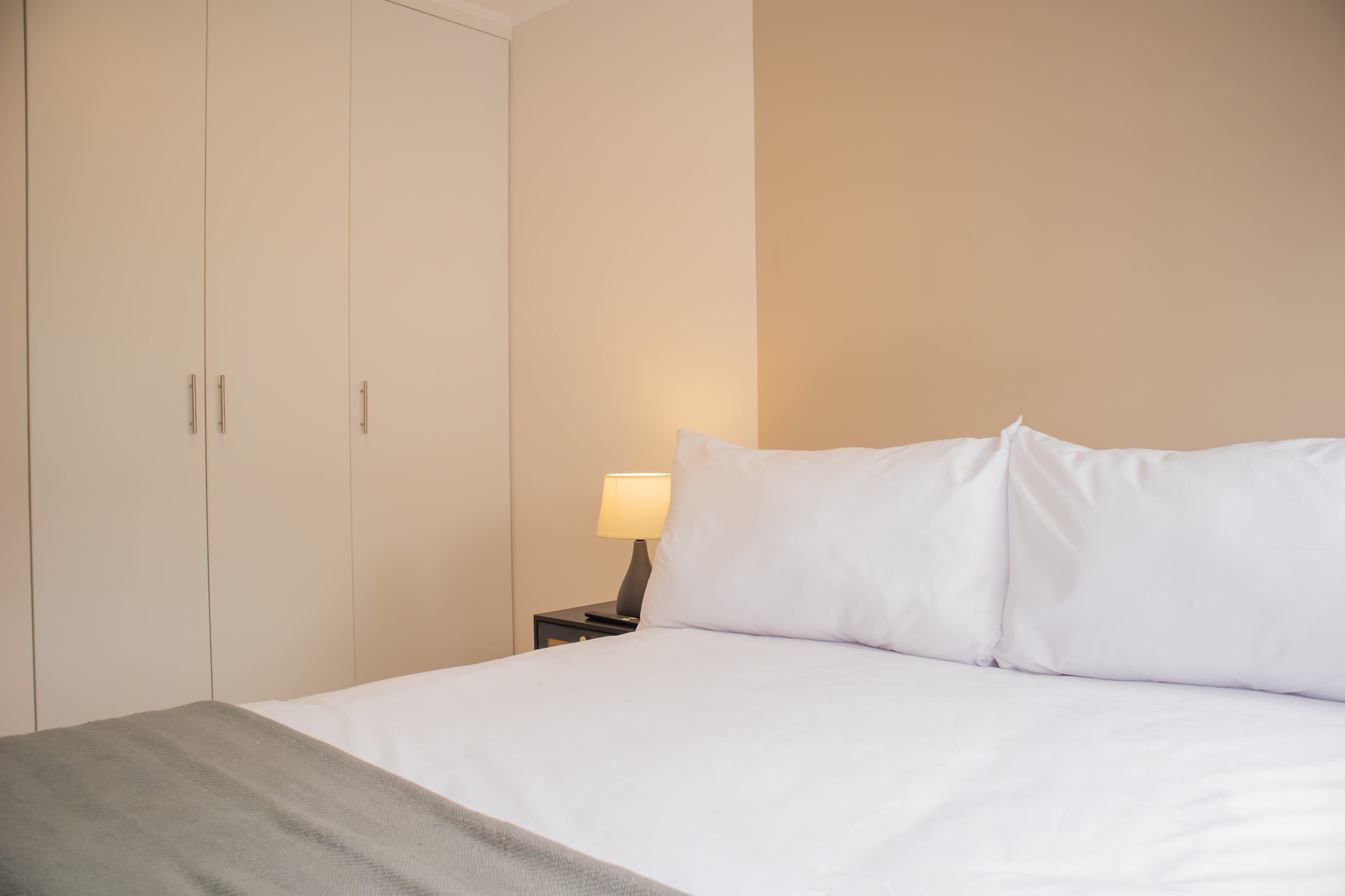 Cama bem arrumada com lençóis brancos, luminária de cabeceira e guarda-roupa com duas portas.