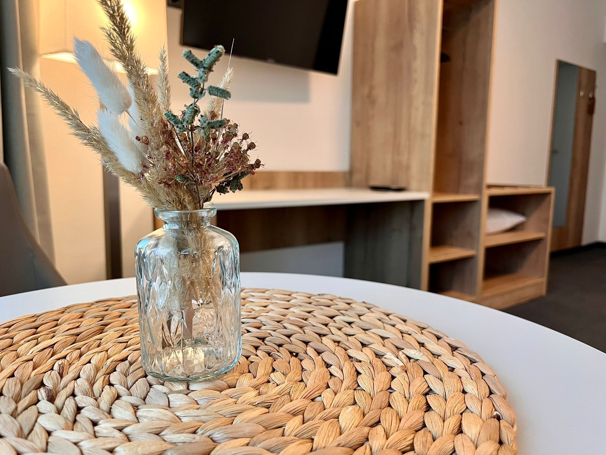 Visão clara de um quarto com um jogo de tabuleiro tecido sobre uma mesa redonda, um vaso de vidro com plantas secas e um armário de madeira ao fundo.