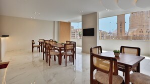 Uma sala de jantar espaçosa com uma mesa grande e cadeiras, uma TV de tela plana na parede e vista para edifícios através de grandes janelas.