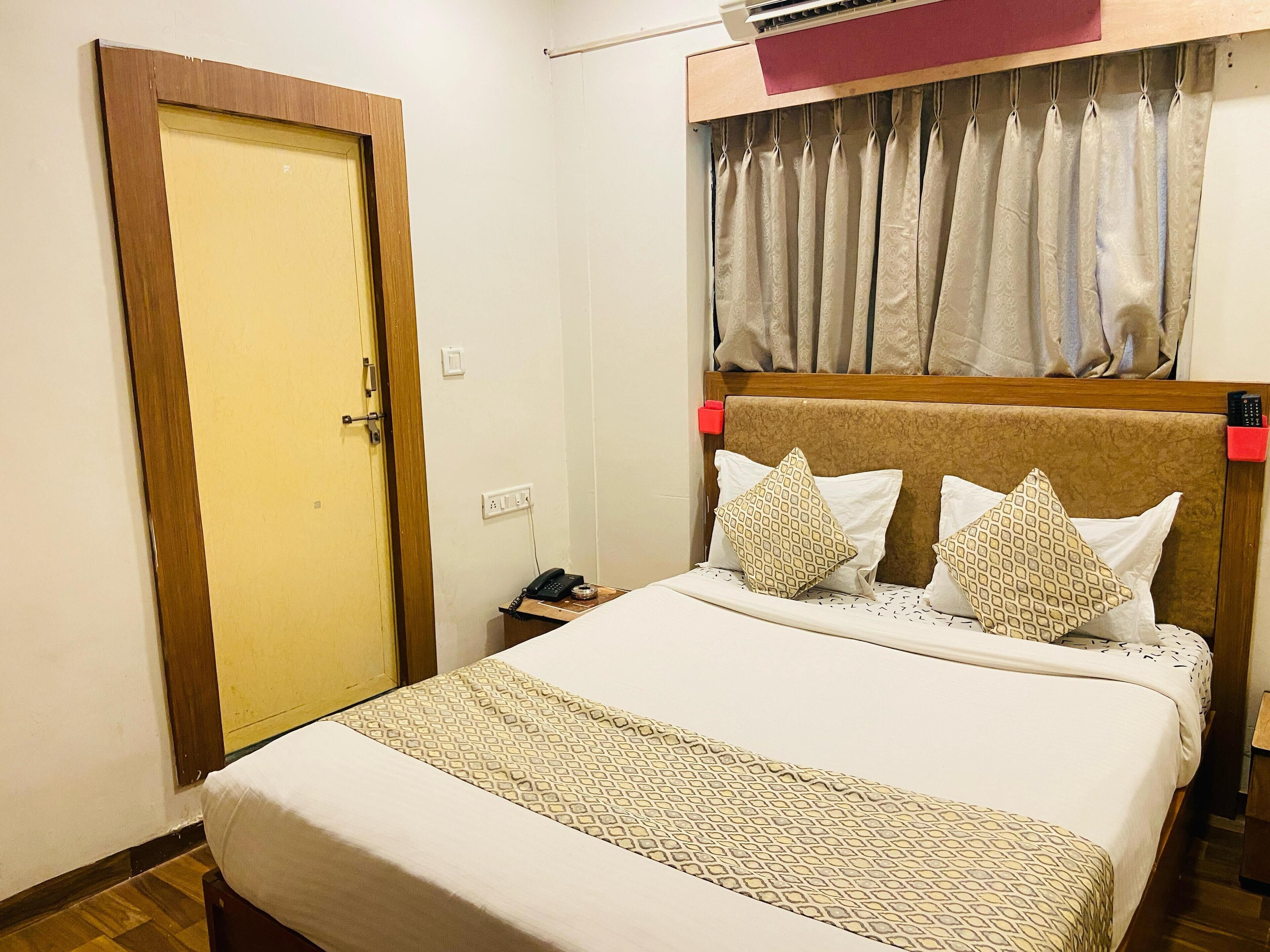 Quarto de hotel com porta de madeira, cama com roupa de cama estampada, telefone de apoio e cortina.