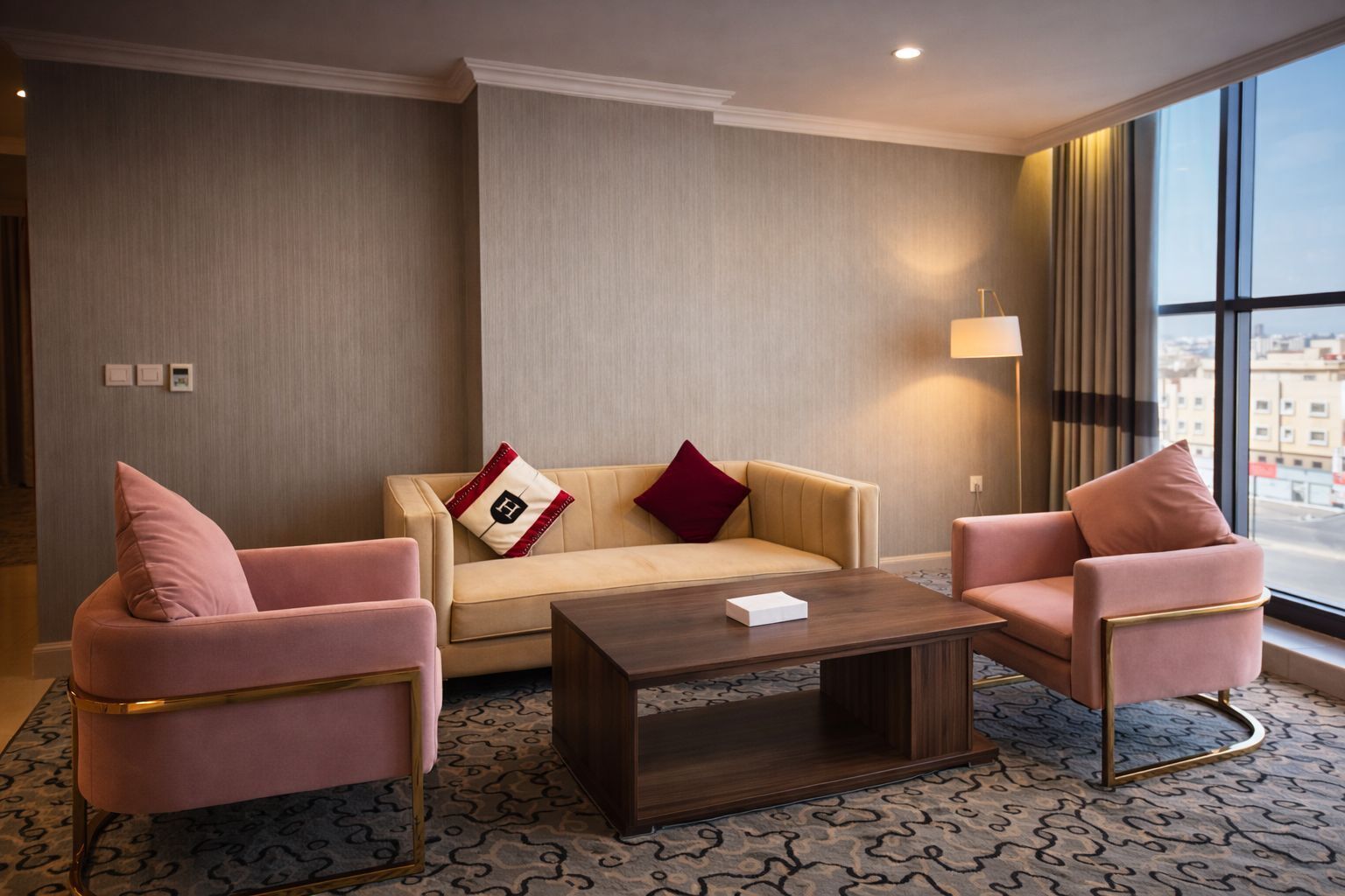 Quarto de hotel com sofá bege, duas poltronas rosa, mesa de centro de madeira e uma janela grande com vista para a cidade.