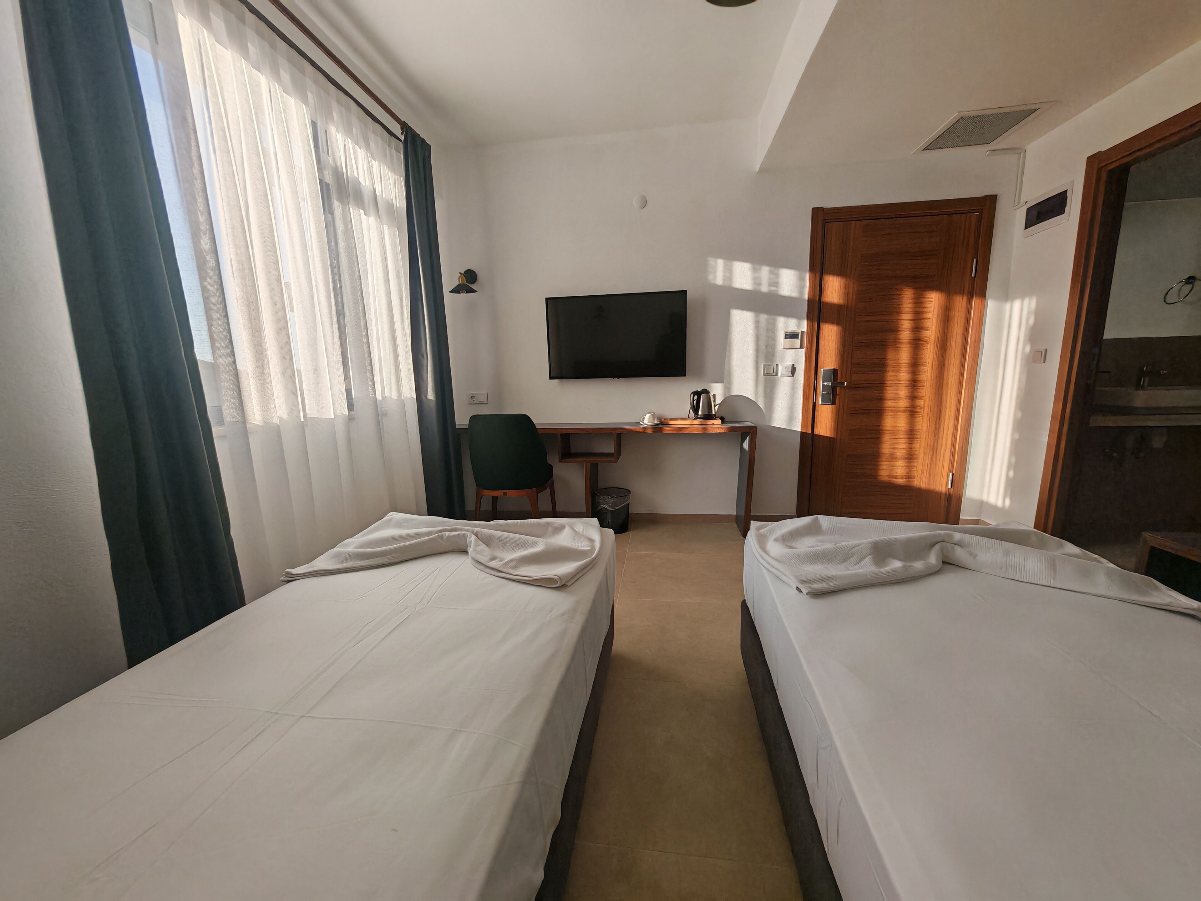 Quarto de hotel com duas camas de solteiro, uma escrivaninha com cadeira, uma televisão e uma porta de madeira.