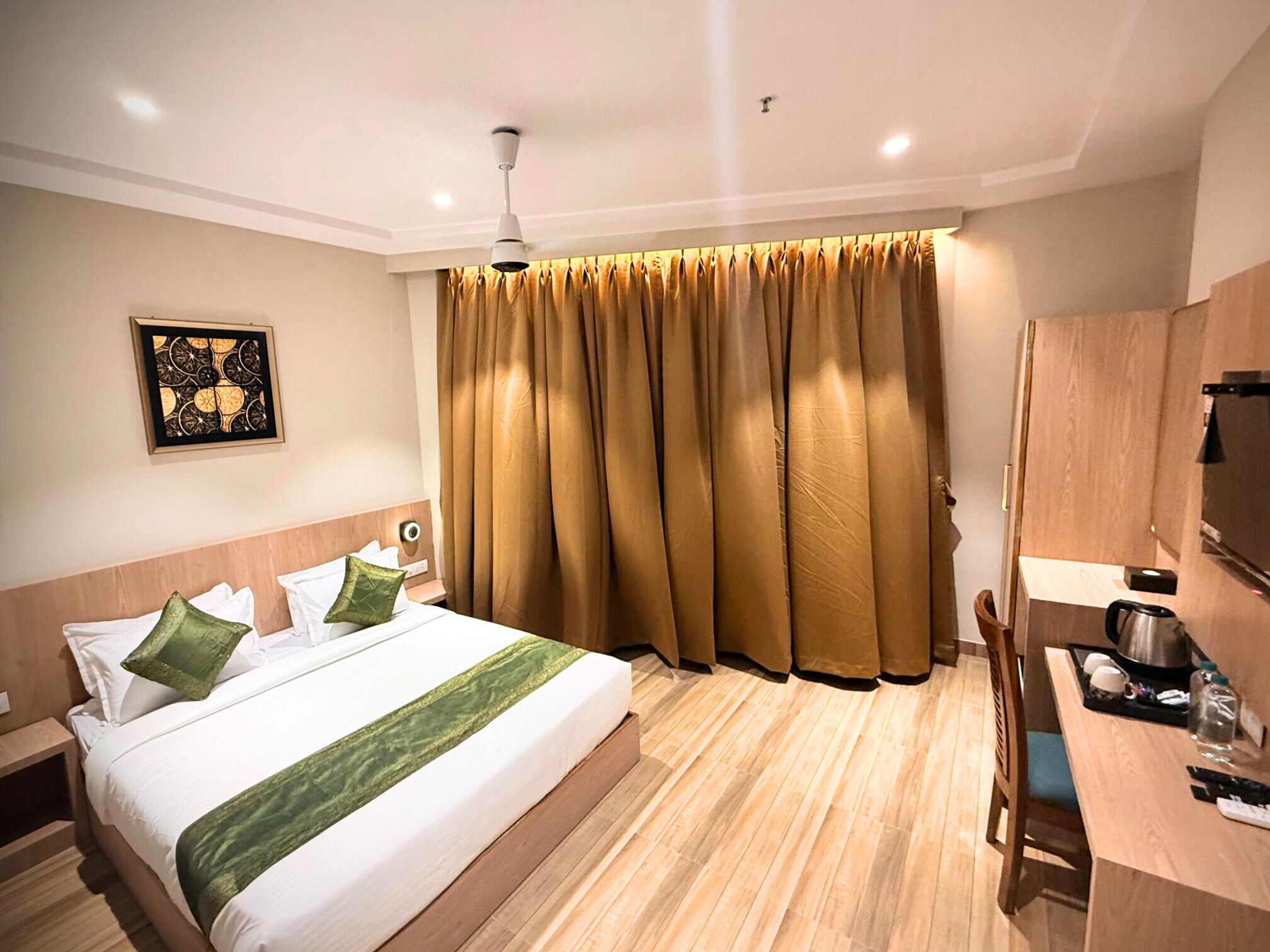 Quarto de hotel com cama, cortinas, um quadro, uma escrivaninha com chaleira e uma cadeira.