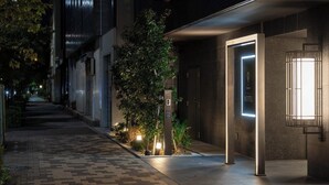 Vista noturna da entrada de um hotel com design moderno, um caminho de paralelepípedos e sinalização iluminada.