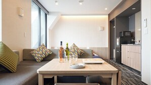 Quarto de hotel moderno com área de refeições, um sofá com almofadas estampadas, uma mesa com garrafas de vinho e copos, e uma cozinha com geladeira e micro-ondas.