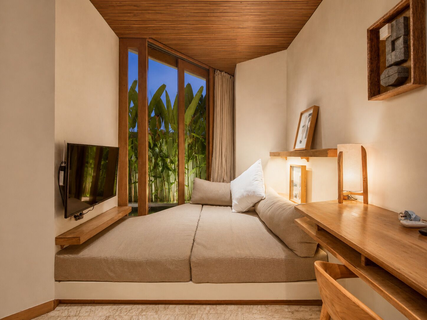 Quarto com cama, televisão, escrivaninha de madeira e vista para o verde.