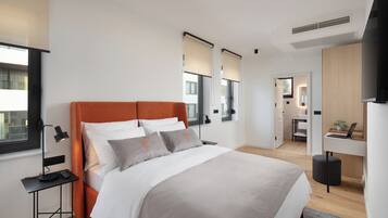 Quarto de hotel moderno com uma cama grande, uma mesa com um laptop, um espelho e uma janela com persianas.