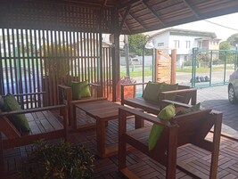 Uma área de estar externa com móveis de madeira e almofadas verdes, uma estrutura coberta e vista para um portão.