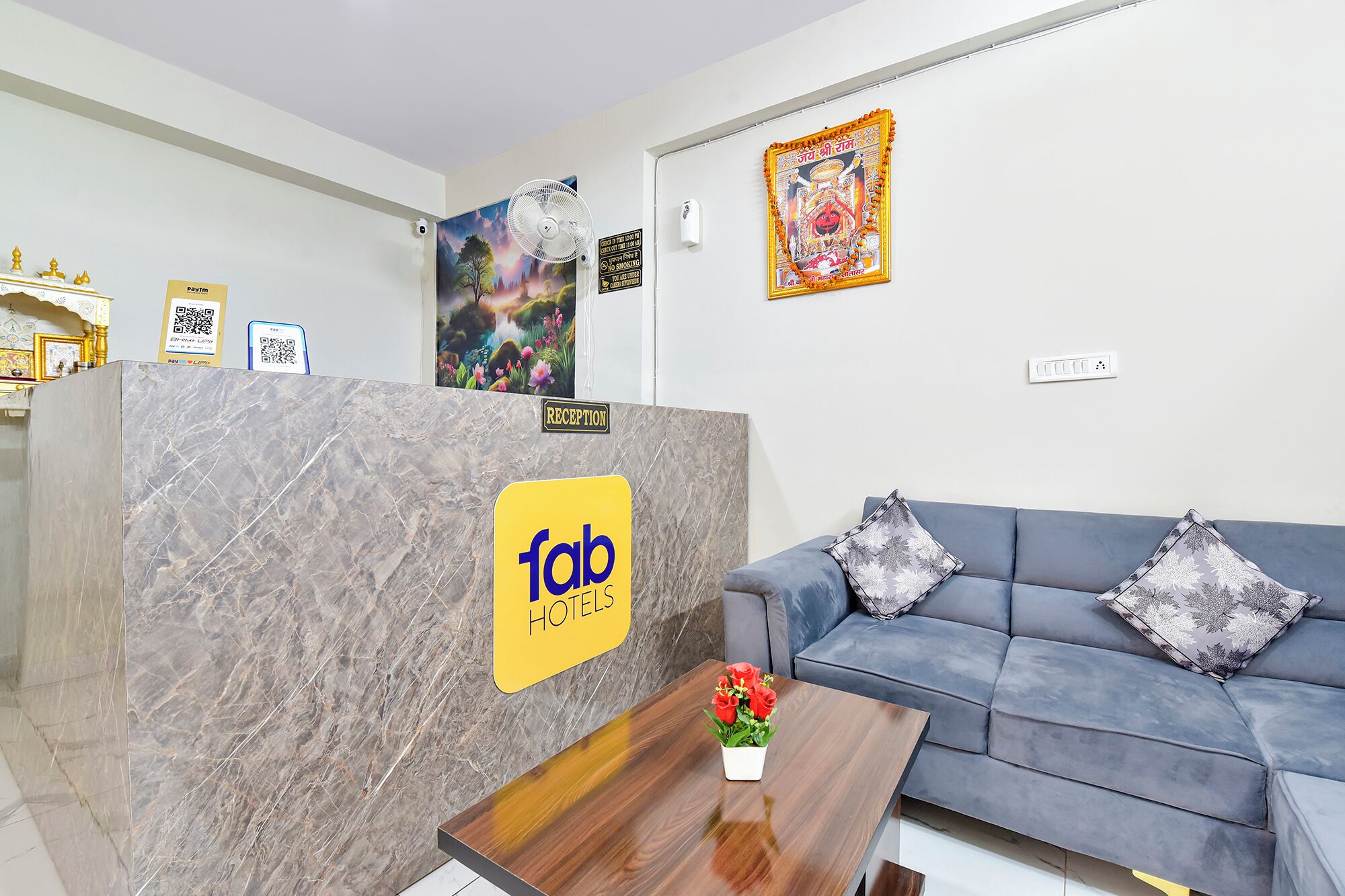 Uma área de recepção de hotel com um balcão de mármore, um sofá com almofadas florais, e uma parede com uma pintura religiosa e uma moldura com uma fotografia.