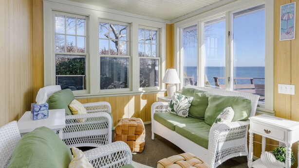 Um quarto iluminado pelo sol, com móveis de vime, janelas amplas e vista para o mar.