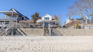 Um imóvel à beira-mar com uma casa de dois andares, uma escadaria que leva a uma porta azul e uma praia arenosa em primeiro plano.