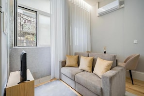 Uma sala de estar moderna com um sofá cinzento, uma televisão de tela plana em um suporte de madeira, e uma janela com cortinas decorativas.