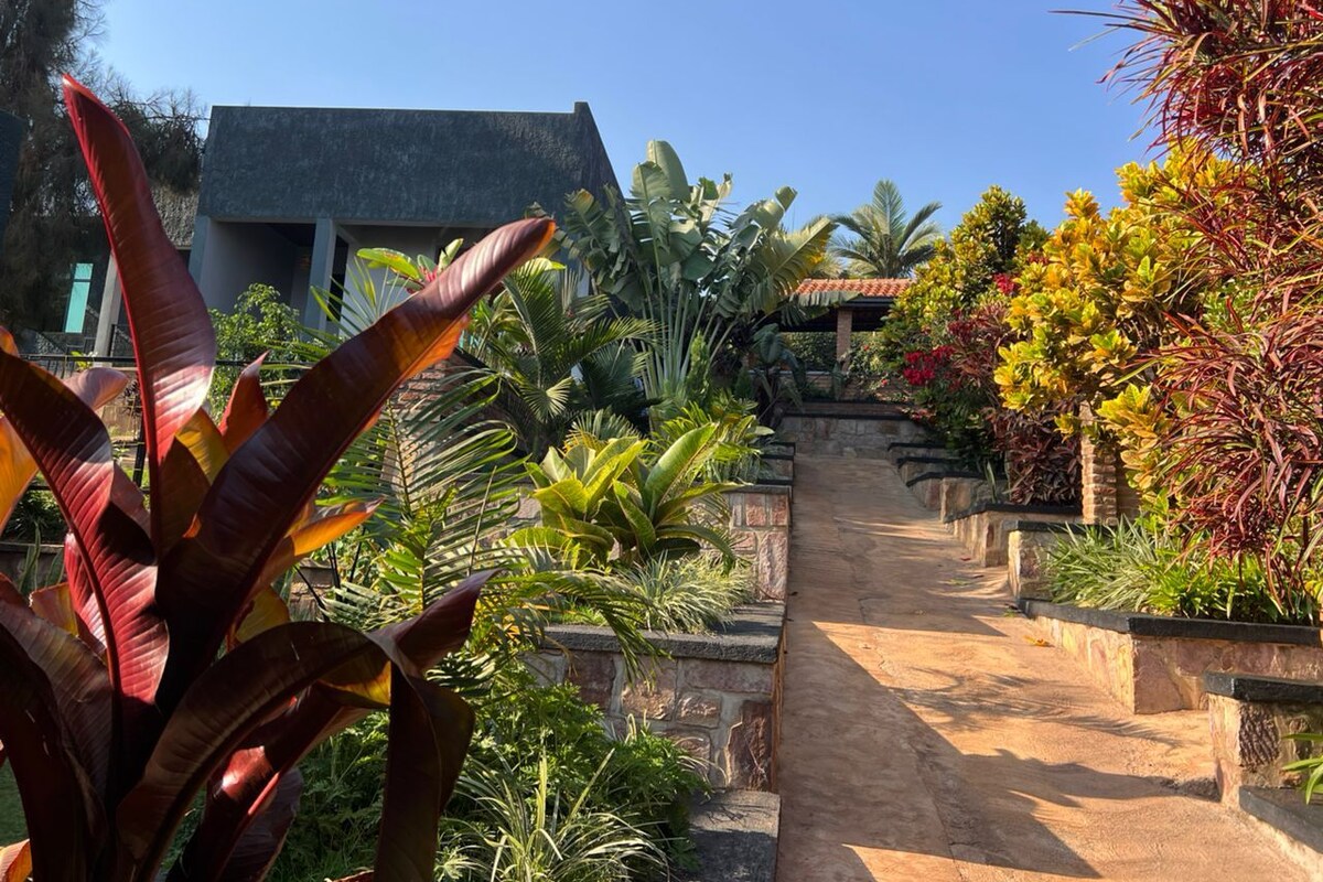 Uma passagem que leva a um edifício com telhado escuro, rodeado por diversas plantas tropicais e uma parede de pedra.