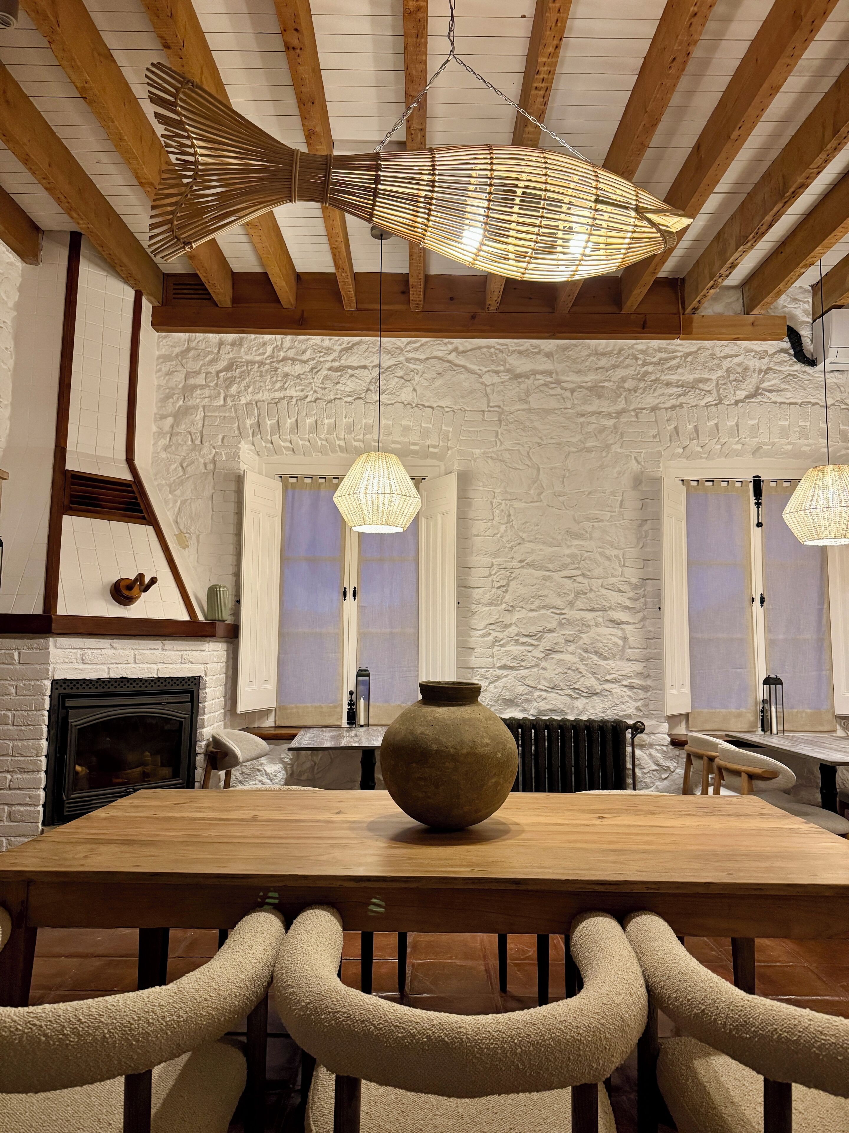 Um interior rústico com vigas de madeira aparentes, uma parede de pedra, uma lareira e duas luminárias pendentes.