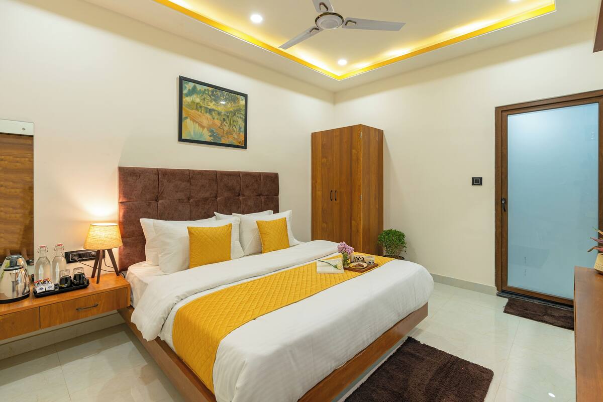 Quarto de hotel com uma cama grande, um guarda-roupa de madeira, uma mesa de cabeceira com abajur e um ventilador de teto.