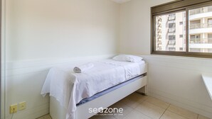 Quarto pequeno e limpo com uma cama de solteiro, janela com vista para um edifício e uma parede branca.