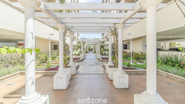 Uma galeria coberta com colunas brancas e piso de azulejos, cercada por vegetação e plantas.