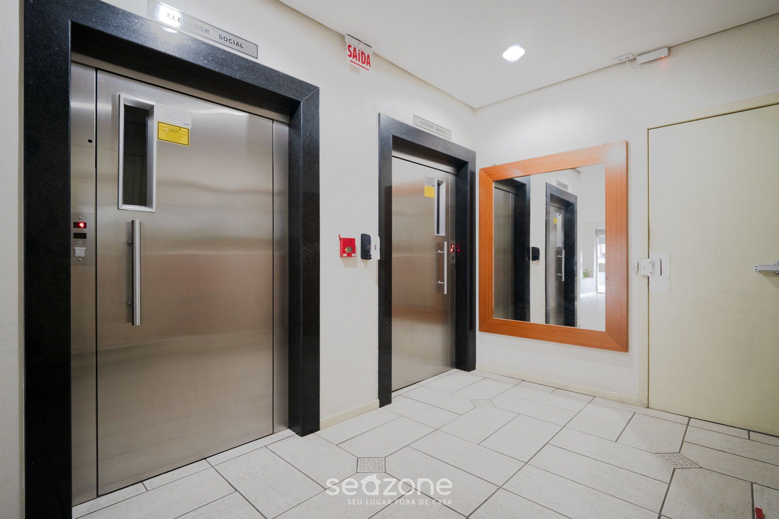 Uma área de elevadores com dois elevadores, uma placa indicando que o elevador está fora de serviço e um espelho na parede.