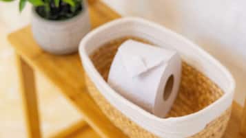 Uma prateleira de madeira com uma cesta de vime contendo um rolo de papel higiênico e uma planta em vaso.