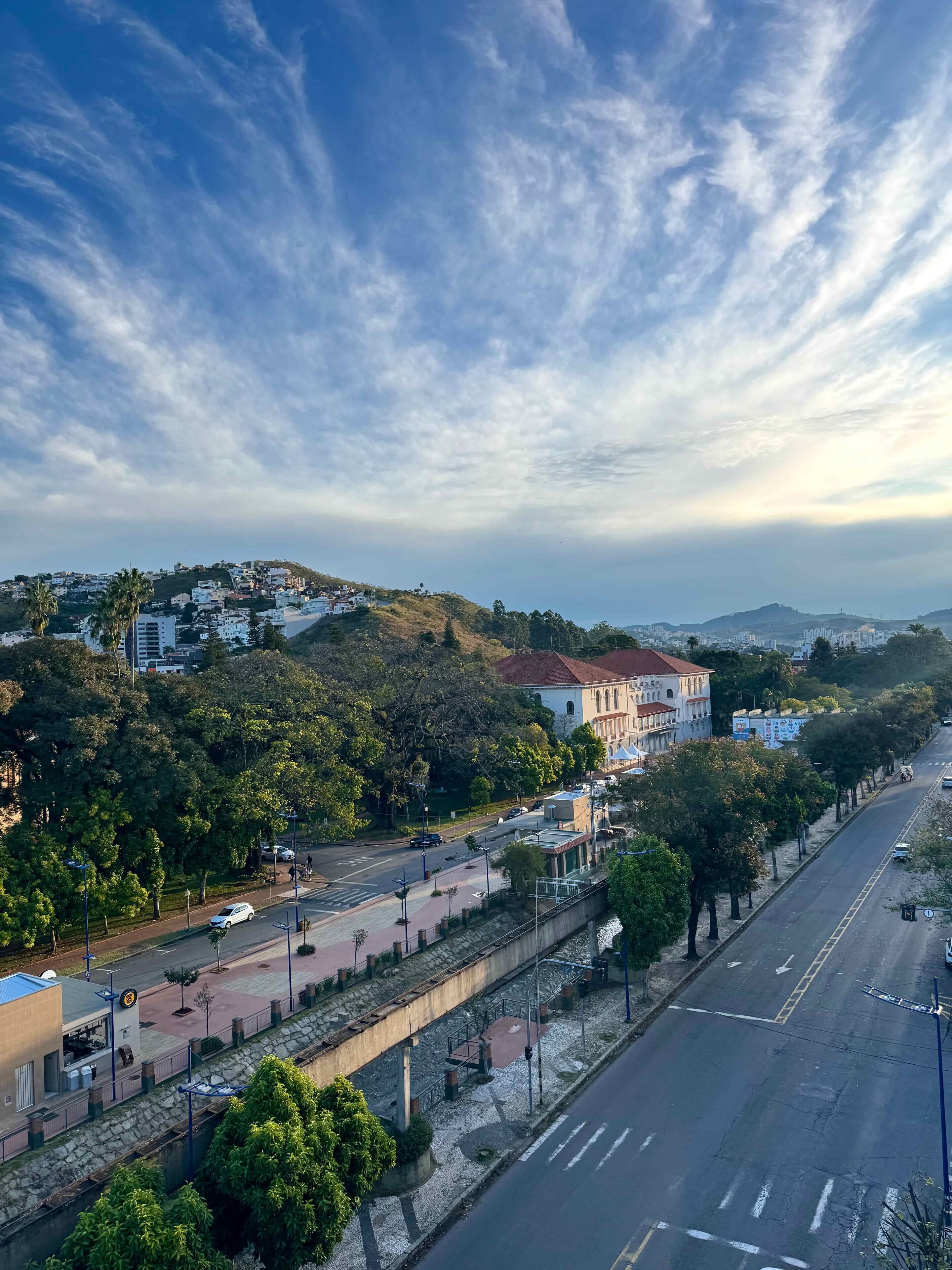 Paisagem costeira urbana com um edifício branco de destaque e telhado vermelho, uma estrada com calçada para pedestres e um corpo d'água.