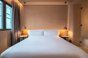 Uma cama bem arrumada em quarto de hotel, com uma janela grande, duas mesas de cabeceira com abajures e um telefone fixado na parede.
