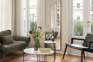 Uma sala de estar com um sofá, uma poltrona, uma mesinha com um vaso de flores e uma garrafa de vinho.