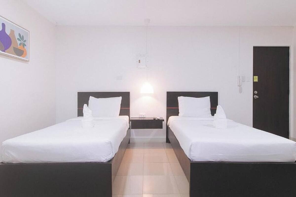 Quarto de hotel com duas camas de solteiro, uma mesa de cabeceira, uma luminária na parede e uma porta.