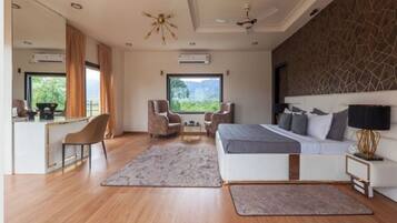 Quarto de hotel moderno com uma cama grande, uma escrivaninha, duas poltronas, um sofá e vista para o verde.