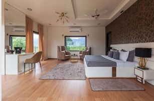 Quarto de hotel moderno com uma cama grande, uma escrivaninha, duas poltronas, um sofá e vista para o verde.