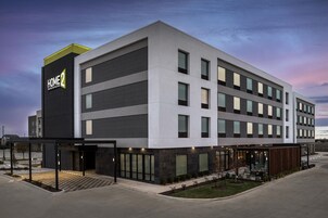 Edifício moderno de hotel com um destaque para a placa 'HOME 2', várias janelas e uma área de entrada bem iluminada.