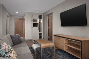 Quarto de hotel moderno com sofá cinzento, mesa de apoio de madeira, televisão fixada na parede e guarda-roupa com portas abertas.