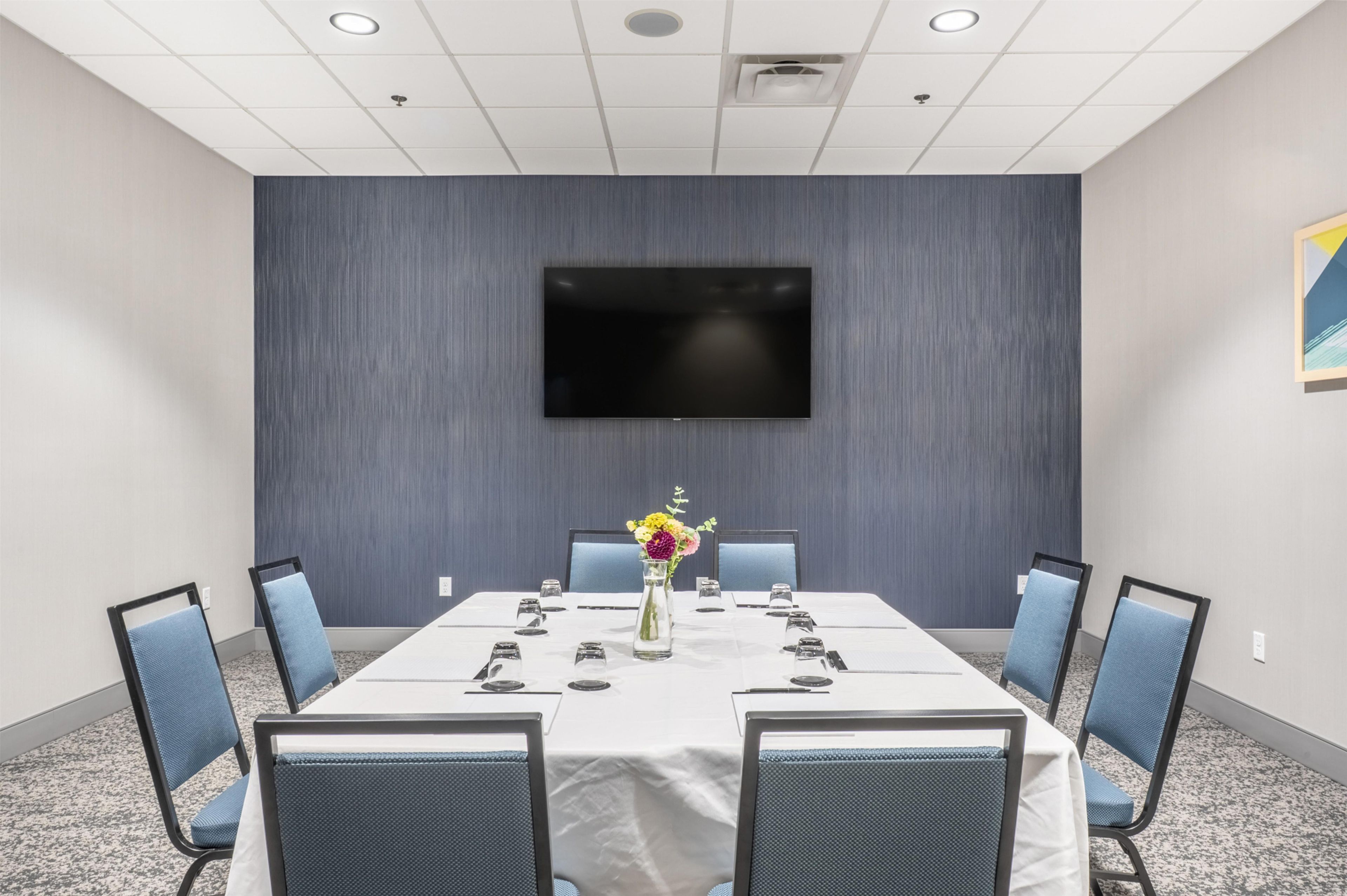 Uma sala de conferências com uma grande televisão de tela plana presa na parede, uma mesa branca com um vaso de flores e seis cadeiras com estofamento azul.