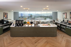 Um café moderno com um balcão central, diversas máquinas de café e vista para a paisagem urbana através de amplas janelas.