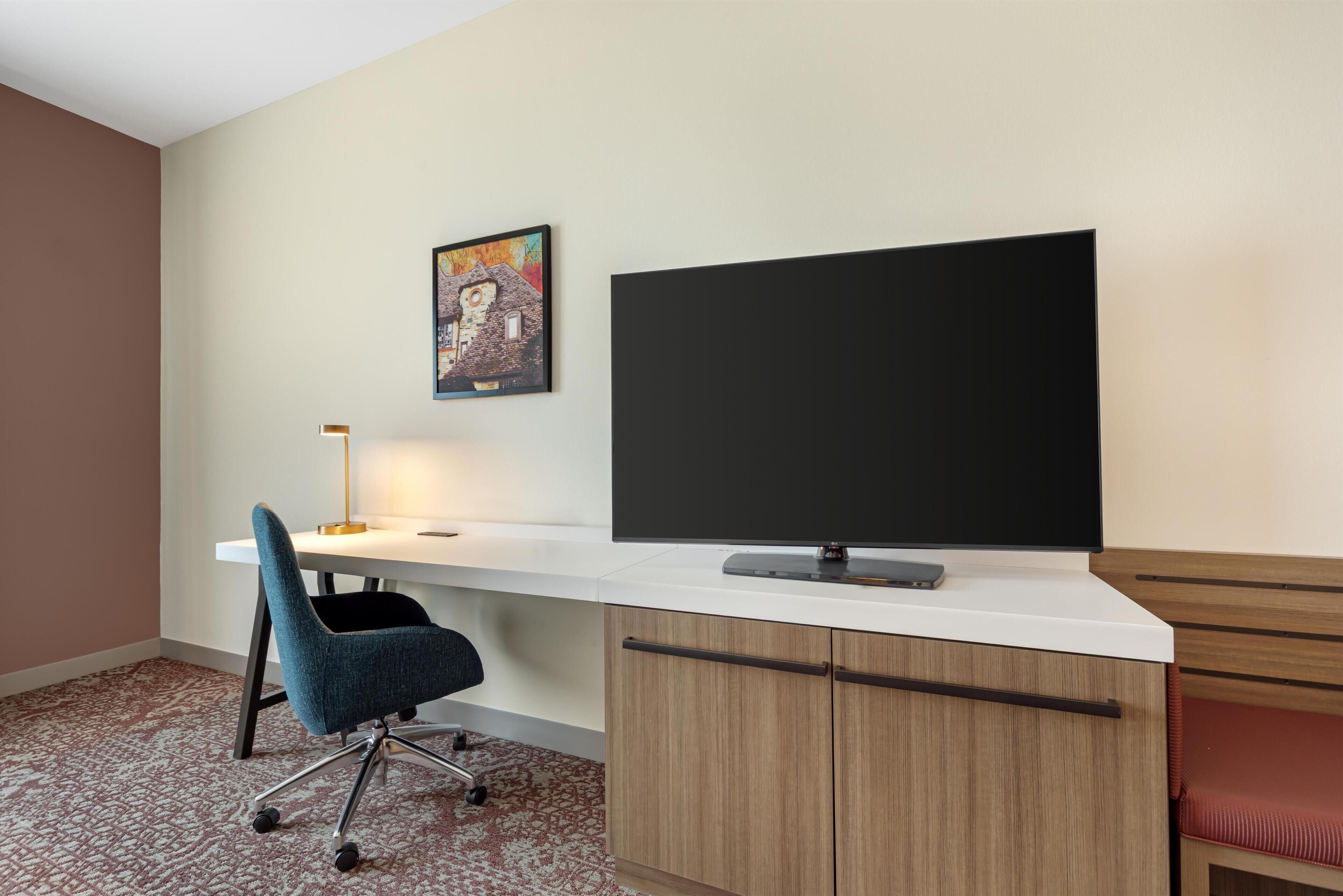 Quarto moderno com escrivaninha branca, cadeira de escritório azul, televisão de tela plana, armário de madeira e uma cadeira vermelha.
