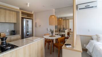 Cozinha moderna com armários de madeira, frigorífico embutido e máquina de café. Existe uma área de refeições com uma mesa redonda e cadeiras, e uma cama com roupa de cama branca no quarto adjacente.