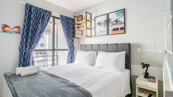 Quarto de hotel com cama, mesa de cabeceira com abajur e janela com cortinas azuis.