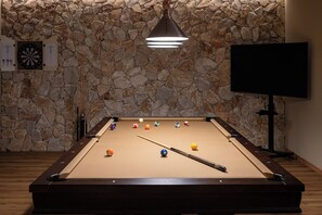 Mesa de bilhar com bolas, alvo de dardos e televisão num espaço com paredes de pedra.