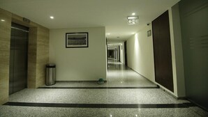 Corredor de hotel com piso de mármore, uma lixeira, um quadro na parede e um elevador à esquerda.