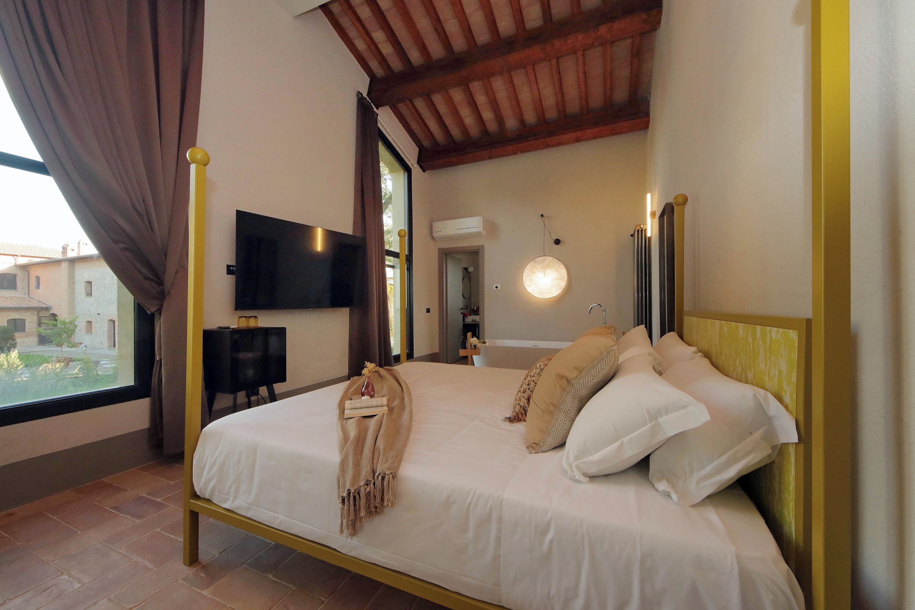 Quarto com cama, televisão, mesa de cabeceira, abajur e janela com cortinas.