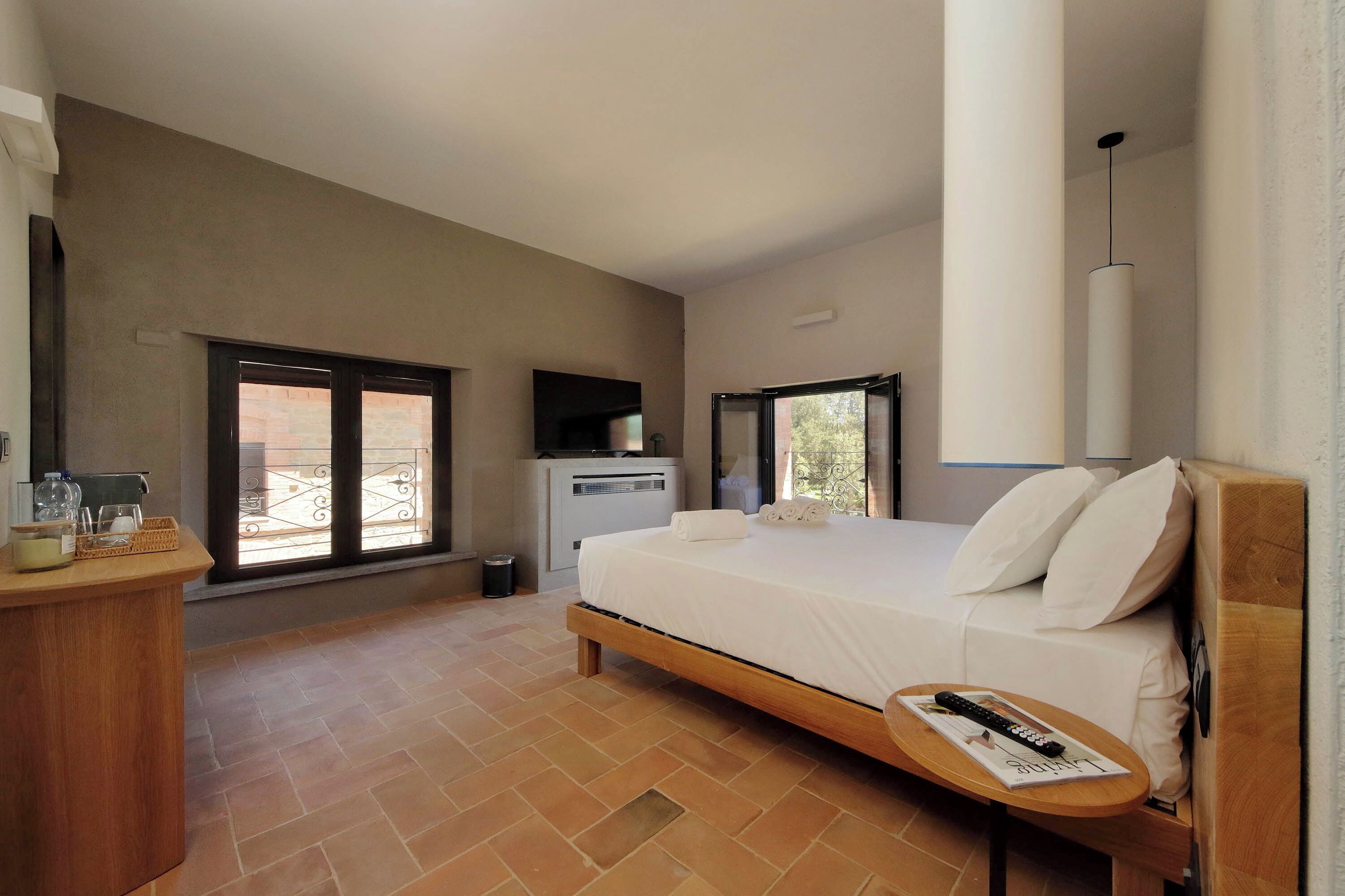 Quarto com cama, mesa de cabeceira de madeira, escrivaninha com cadeira, televisão fixada na parede e janela com vista para o exterior.