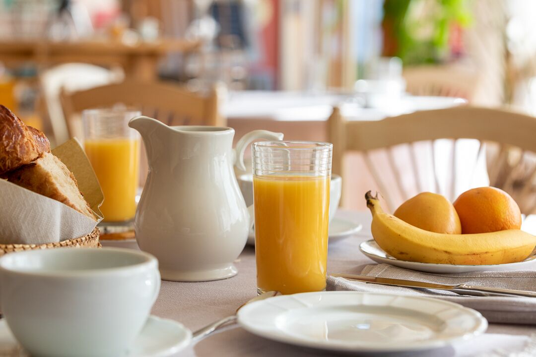 Mesa de café da manhã com pão, jarra, copo de suco de laranja e prato com laranjas e uma banana.