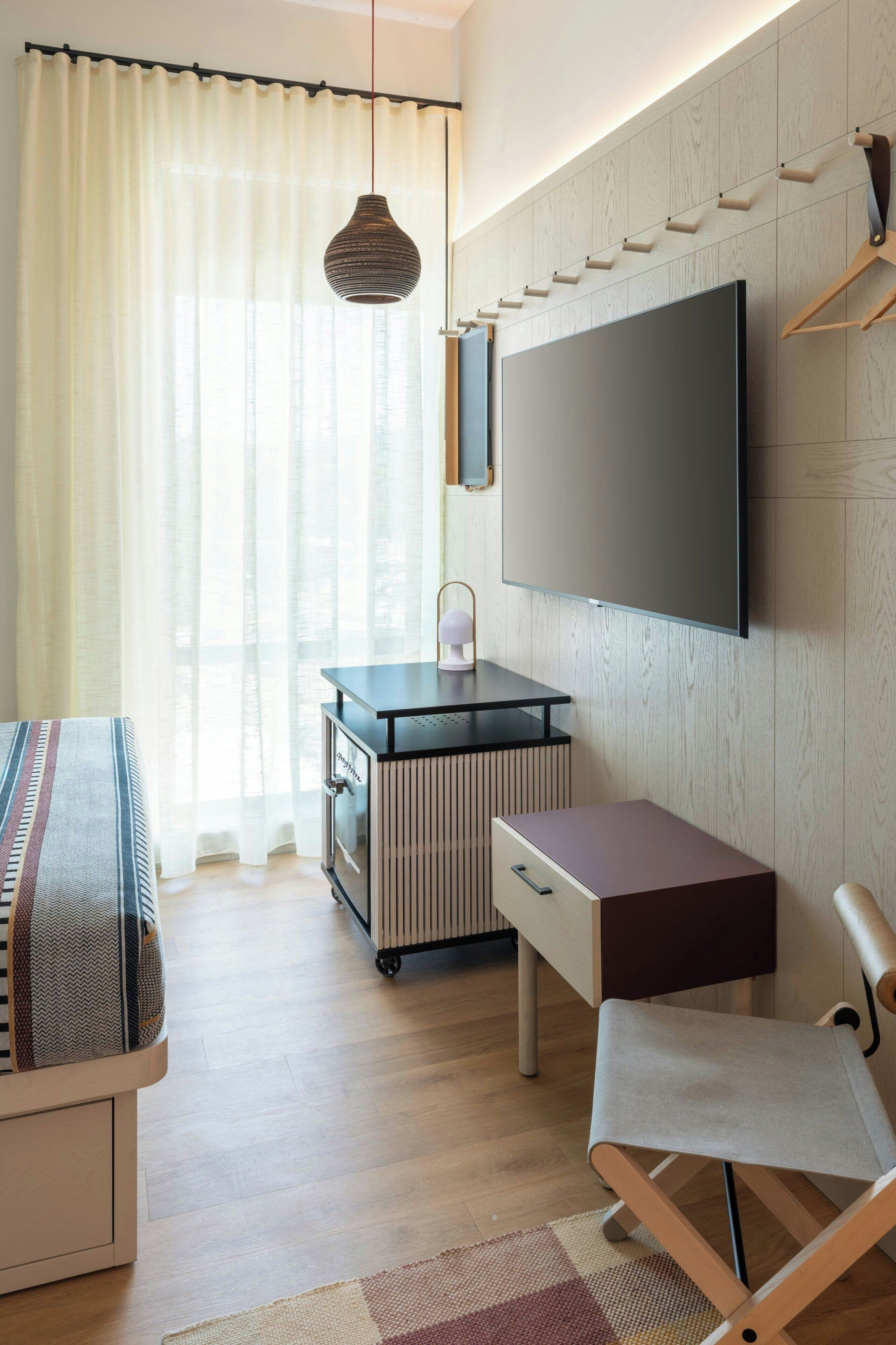 Quarto de hotel moderno com uma televisão de tela grande fixada na parede, um armário de madeira, uma escrivaninha com cadeira e uma cama com colcha estampada.