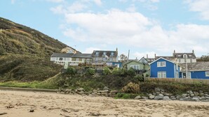 Um complexo de edifícios à beira-mar com várias casas, uma praia de areia em primeiro plano e um cenário de colinas ao fundo.