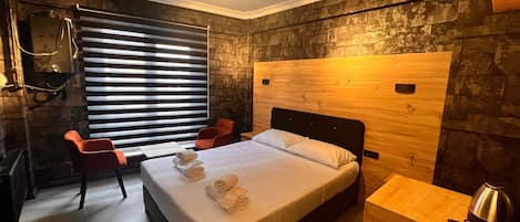 Quarto de hotel moderno com uma cama grande, cabeceira de madeira, uma cadeira vermelha, uma pequena mesa com uma chaleira e uma parede de pedra.
